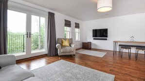 Uma sala de estar ampla, com um sofá, uma poltrona listrada, uma mesa de jantar com cadeiras, uma TV fixada na parede e grandes janelas com cortinas.