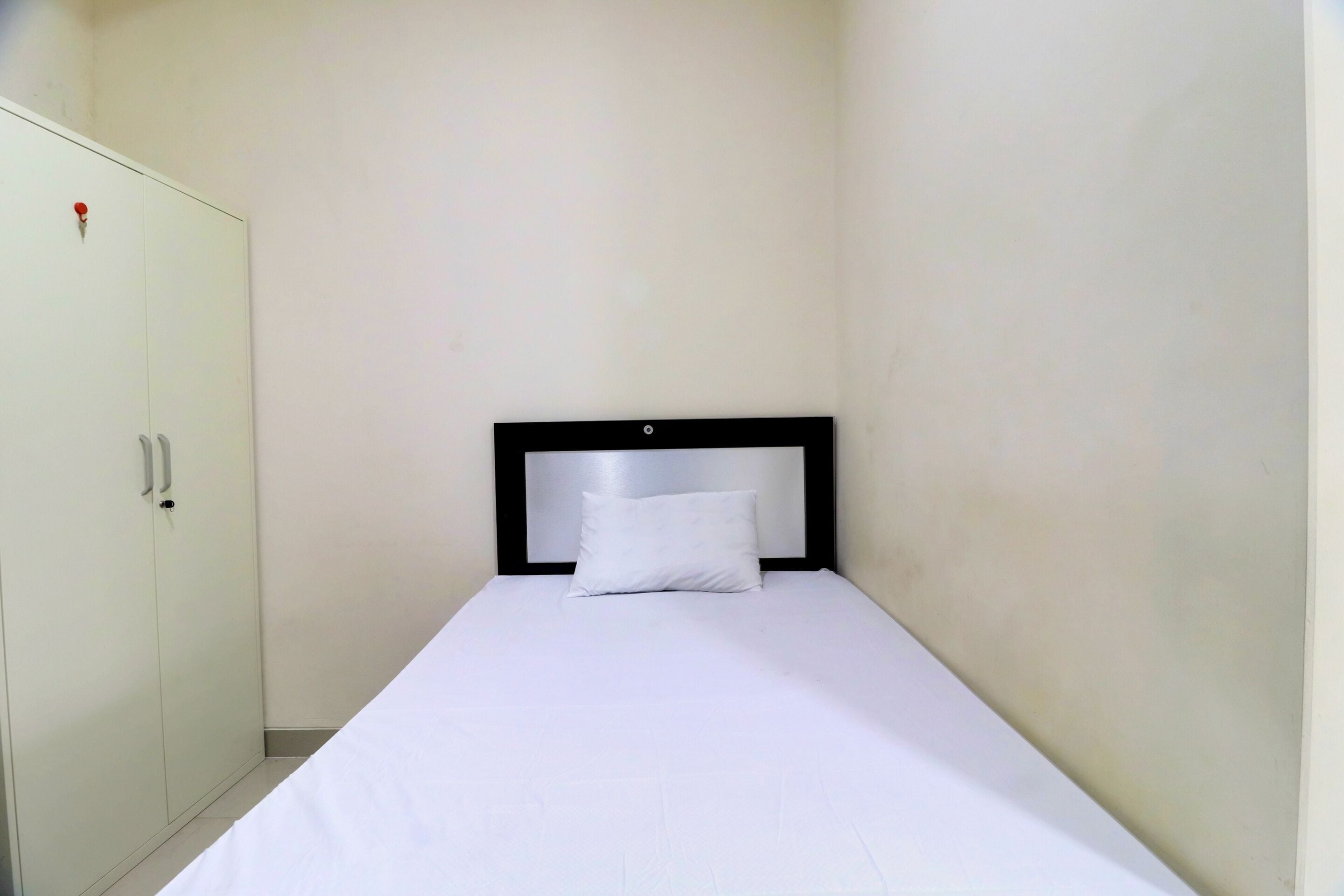 Quarto pequeno com uma cama de solteiro, cabeceira preta e guarda-roupa branco com um objeto vermelho em cima.