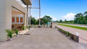 Um edifício moderno com área externa coberta, plantas em vasos e vista para uma estrada e vegetação.