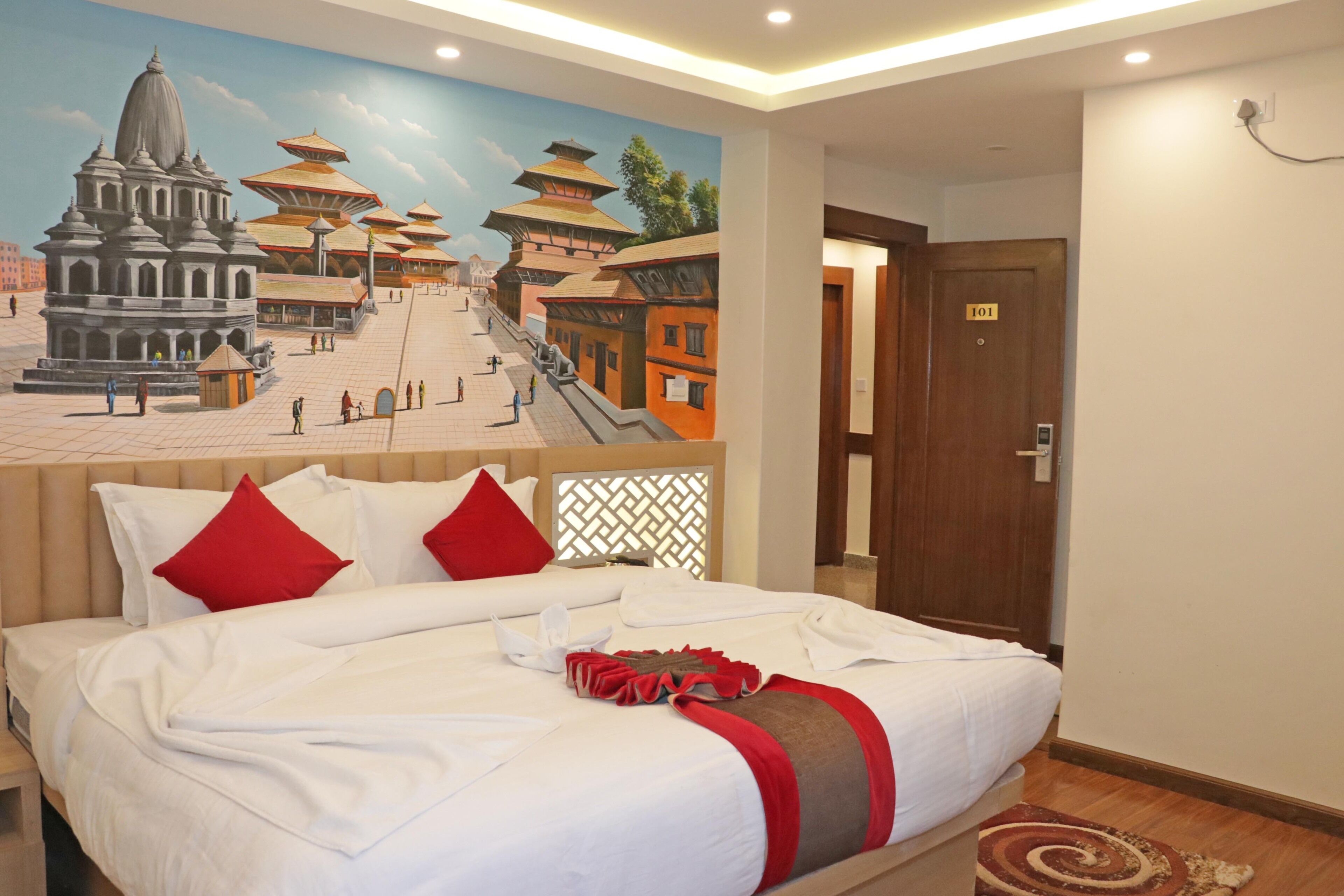 Quarto de hotel com cama grande, almofadas vermelhas, um mural de um templo e uma porta de madeira com a inscrição 101.