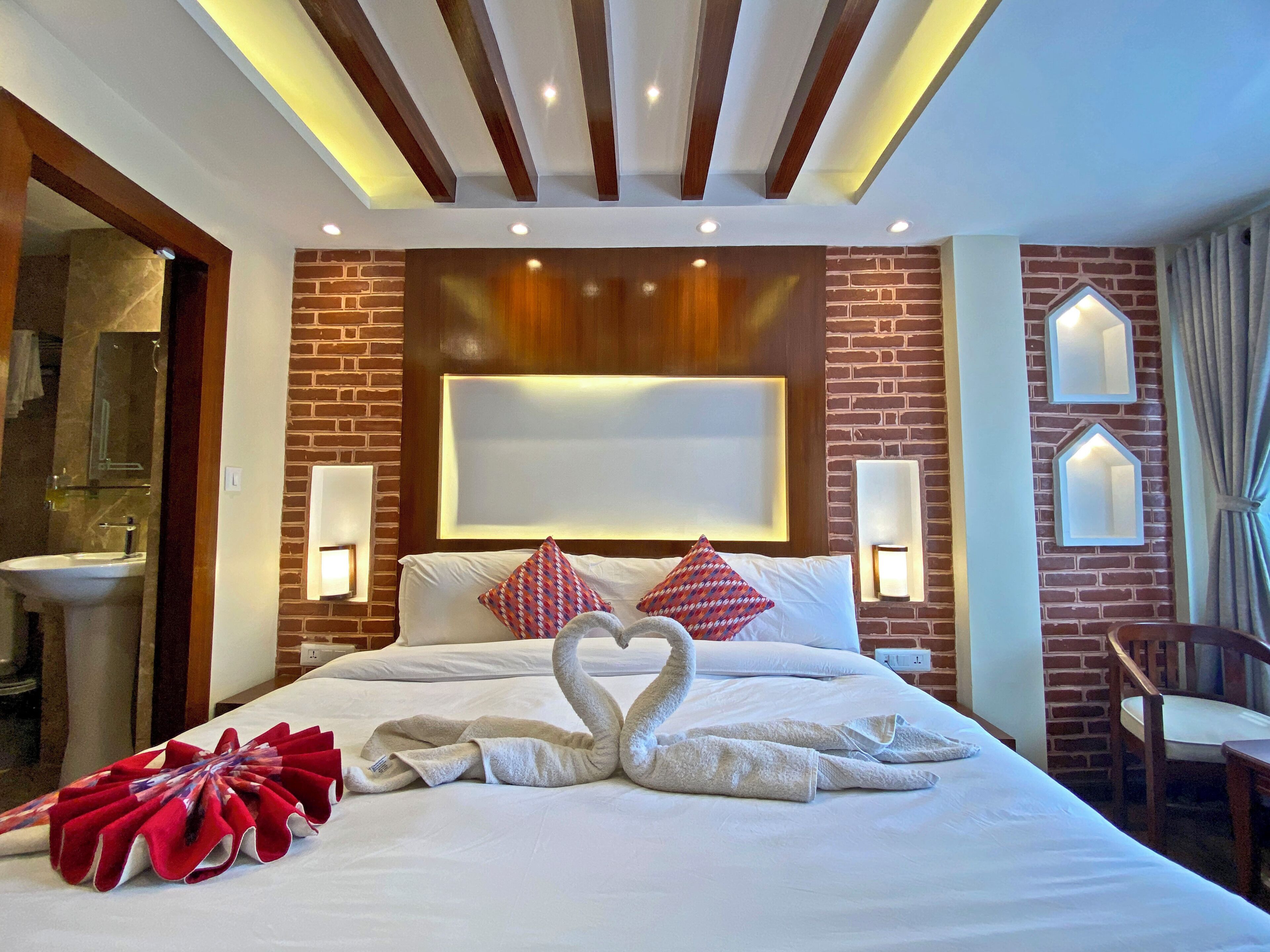 Quarto de hotel com parede de tijolos, uma cama grande com roupa de cama branca, duas luminárias fixadas na parede e teto de madeira.
