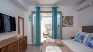 Quarto de hotel com cama, cómoda de madeira, televisão e vista através de uma porta aberta.