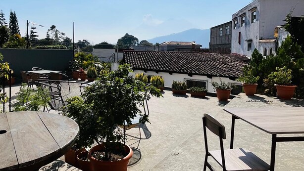 Uma área de terraço ao ar livre com mesas e cadeiras redondas, plantas em vasos e um grande guarda-sol para sombra.
