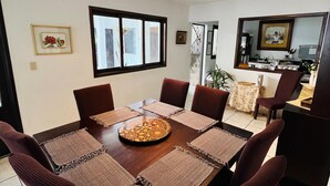 Uma sala de jantar com uma mesa de madeira, seis cadeiras e um objeto decorativo no centro. Há uma janela com vista para o exterior, e uma área de cozinha é visível ao fundo.
