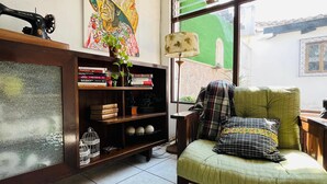 Uma sala de estar com um sofá verde, uma estante de livros, um quadro e uma janela com vista para um edifício verde.