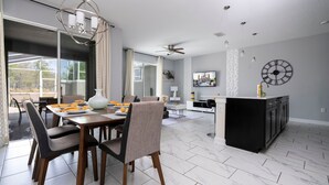 Uma área de refeições moderna com uma mesa grande, cadeiras e um candelabro. Há também uma cozinha com bancada preta e armários.