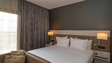 Quarto de hotel com uma cama grande, duas mesas de cabeceira com abajures, um sofá e vista para edifícios pela janela.