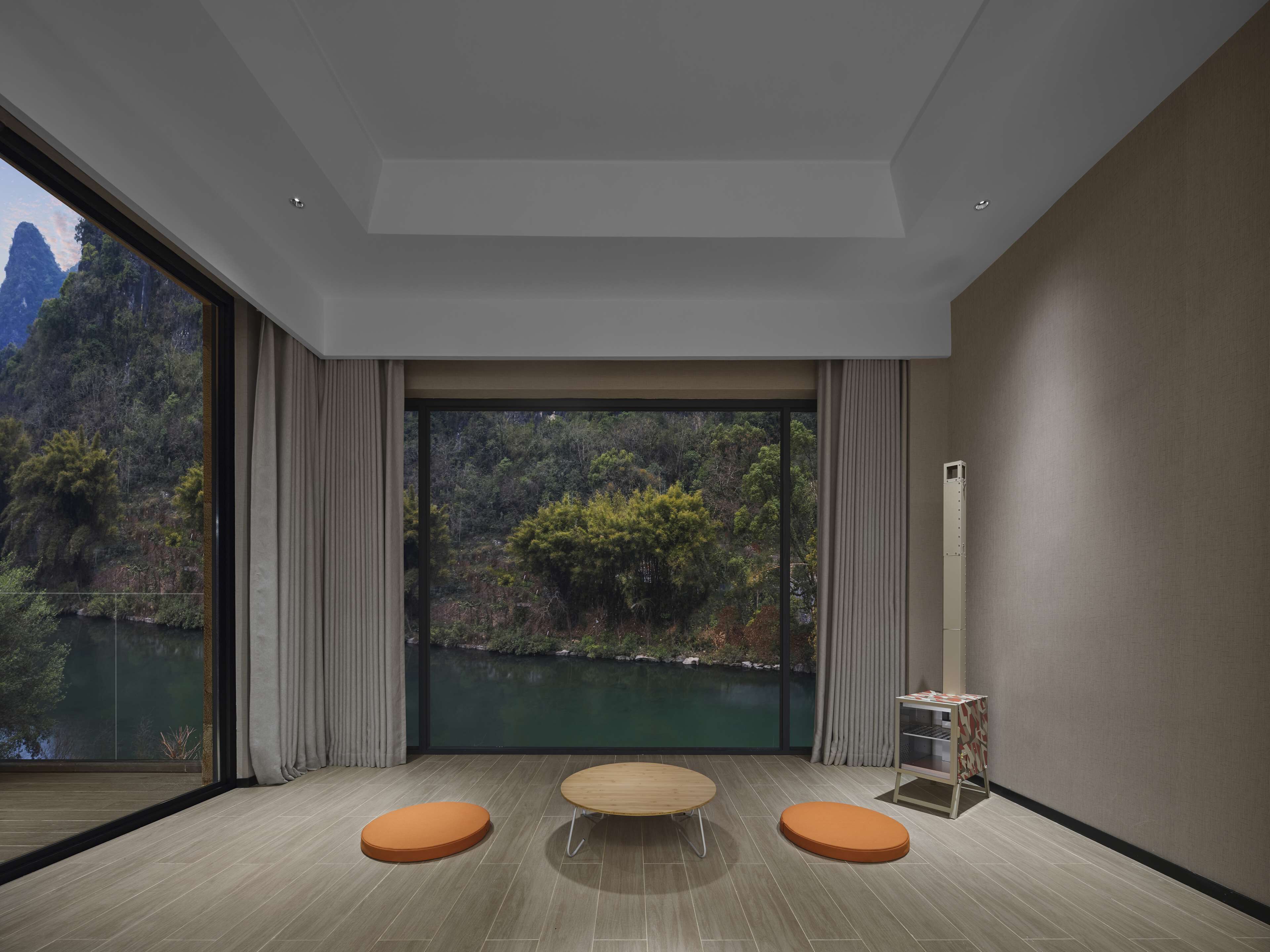 Quarto moderno com uma janela grande, uma mesa redonda e dois banquinhos laranja.
