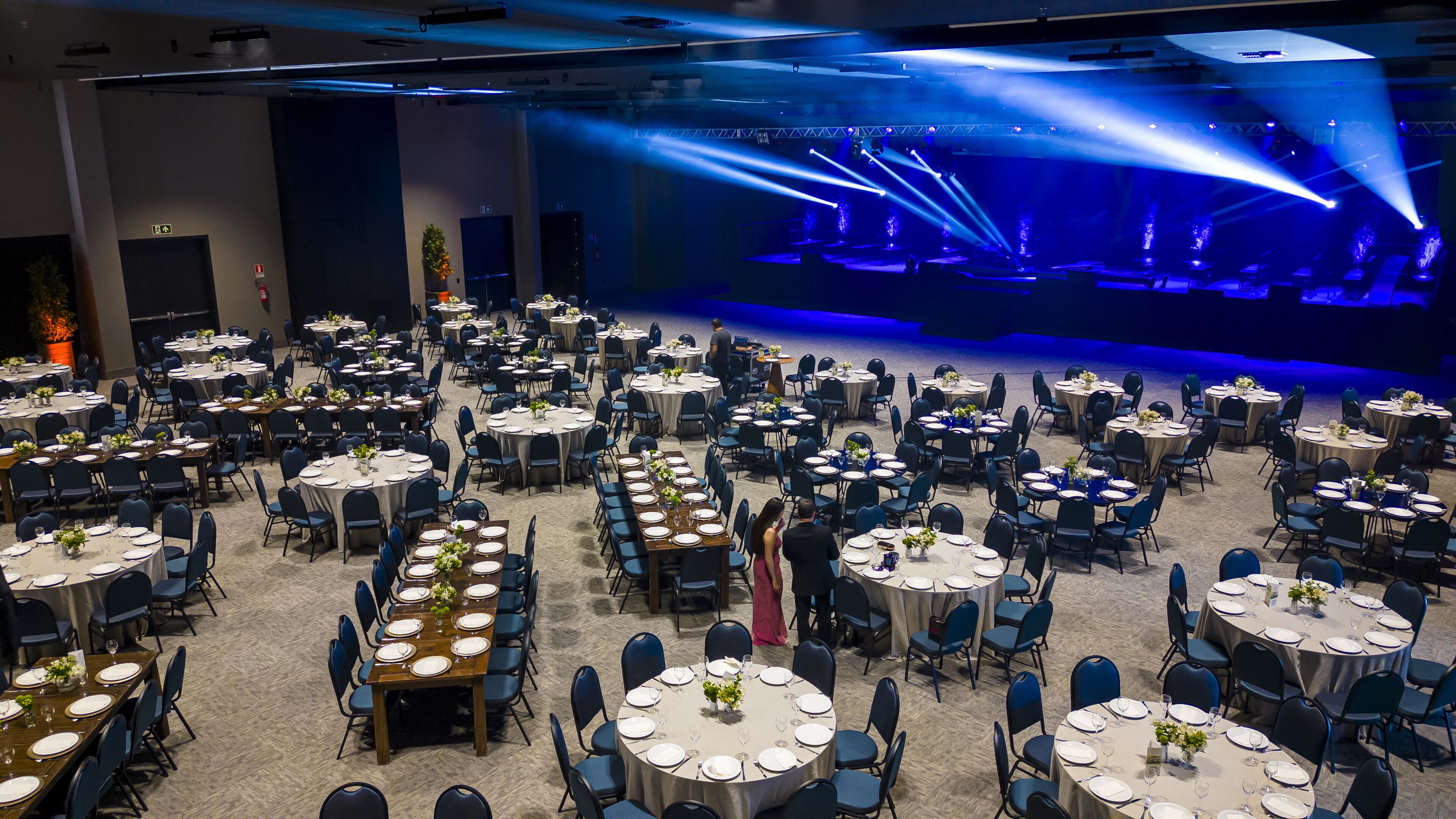 Uma sala de banquetes com mesas redondas preparadas para refeições, um palco com iluminação azul e piso coberto de carpete.