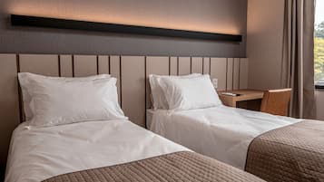 Quarto de hotel com duas camas, uma mesa de cabeceira de madeira e uma janela com cortinas.
