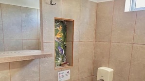 Casa de banho com azulejos bege, sanita, dispensador de sabonete, pequena estante com objeto decorativo e espelho.