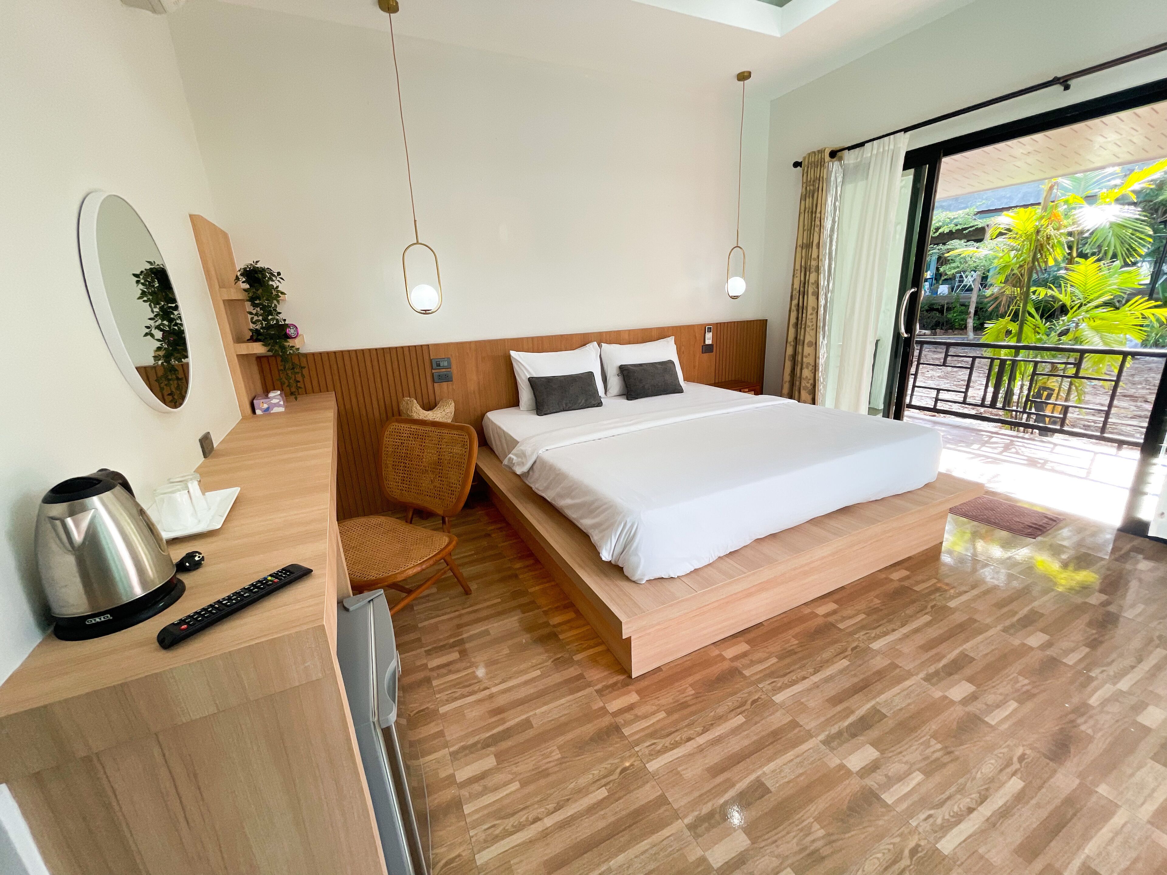 Quarto de hotel moderno com uma cama grande, piso de madeira, escrivaninha com chaleira, uma cadeira e vista para um jardim através de uma porta de correr.
