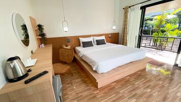 Quarto de hotel moderno com uma cama grande, piso de madeira, escrivaninha com chaleira, uma cadeira e vista para um jardim através de uma porta de correr.