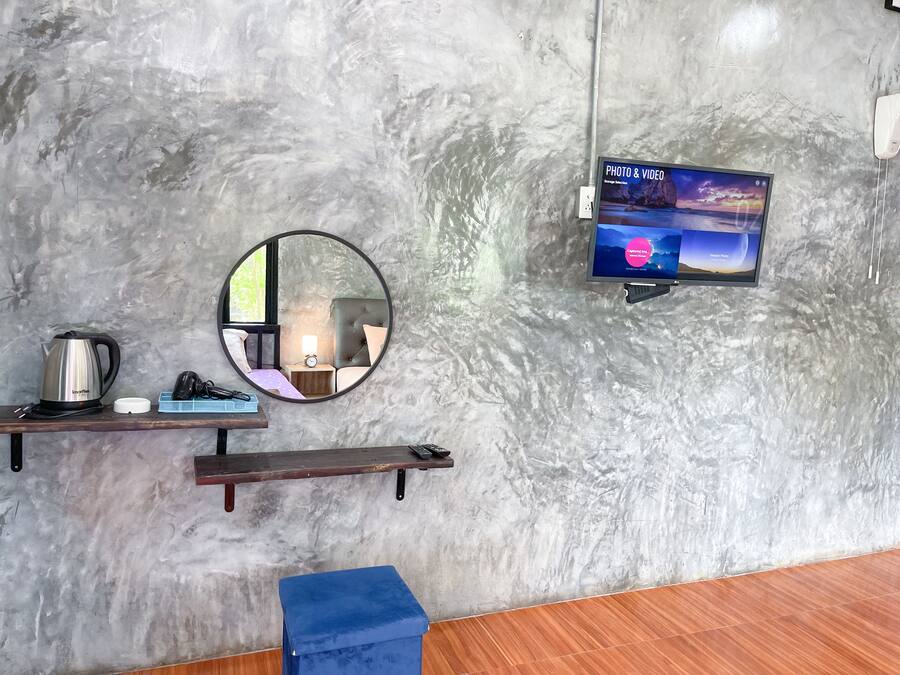 Quarto com parede cinza texturizada, espelho redondo, televisão montada na parede, prateleira de madeira com uma chaleira, luminária e uma caixa azul.