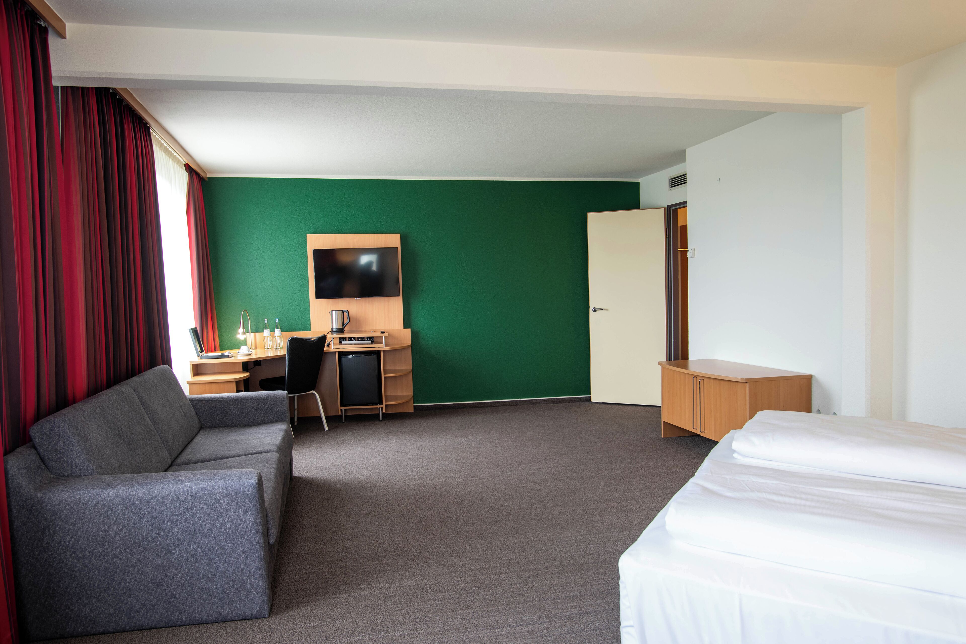 Quarto de hotel com sofá cinzento, uma cama, escrivaninha com computador, televisão e guarda-roupa.