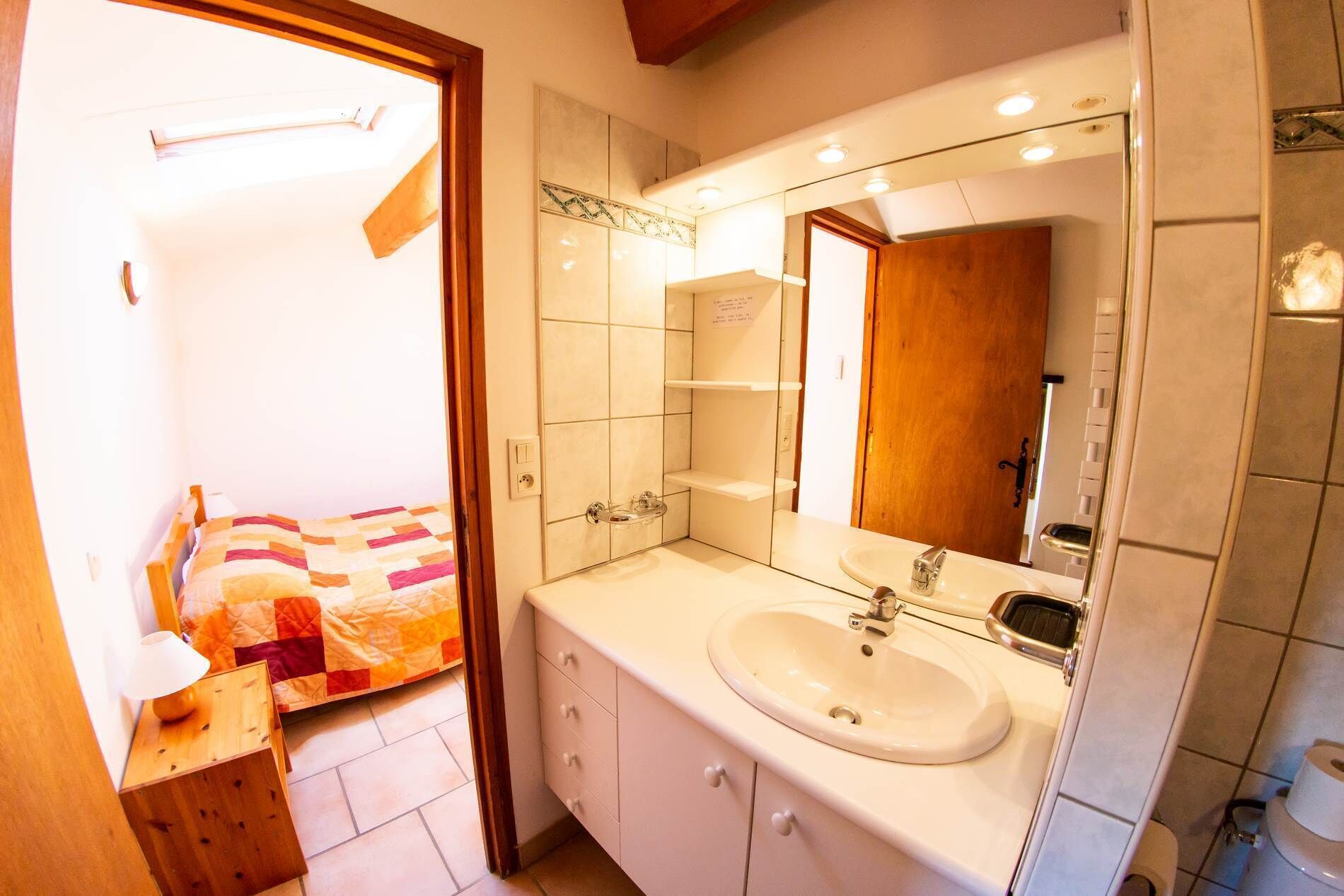 Casa de banho com lavatório, espelho e porta de madeira que dá para um quarto com cama e mesa de cabeceira.