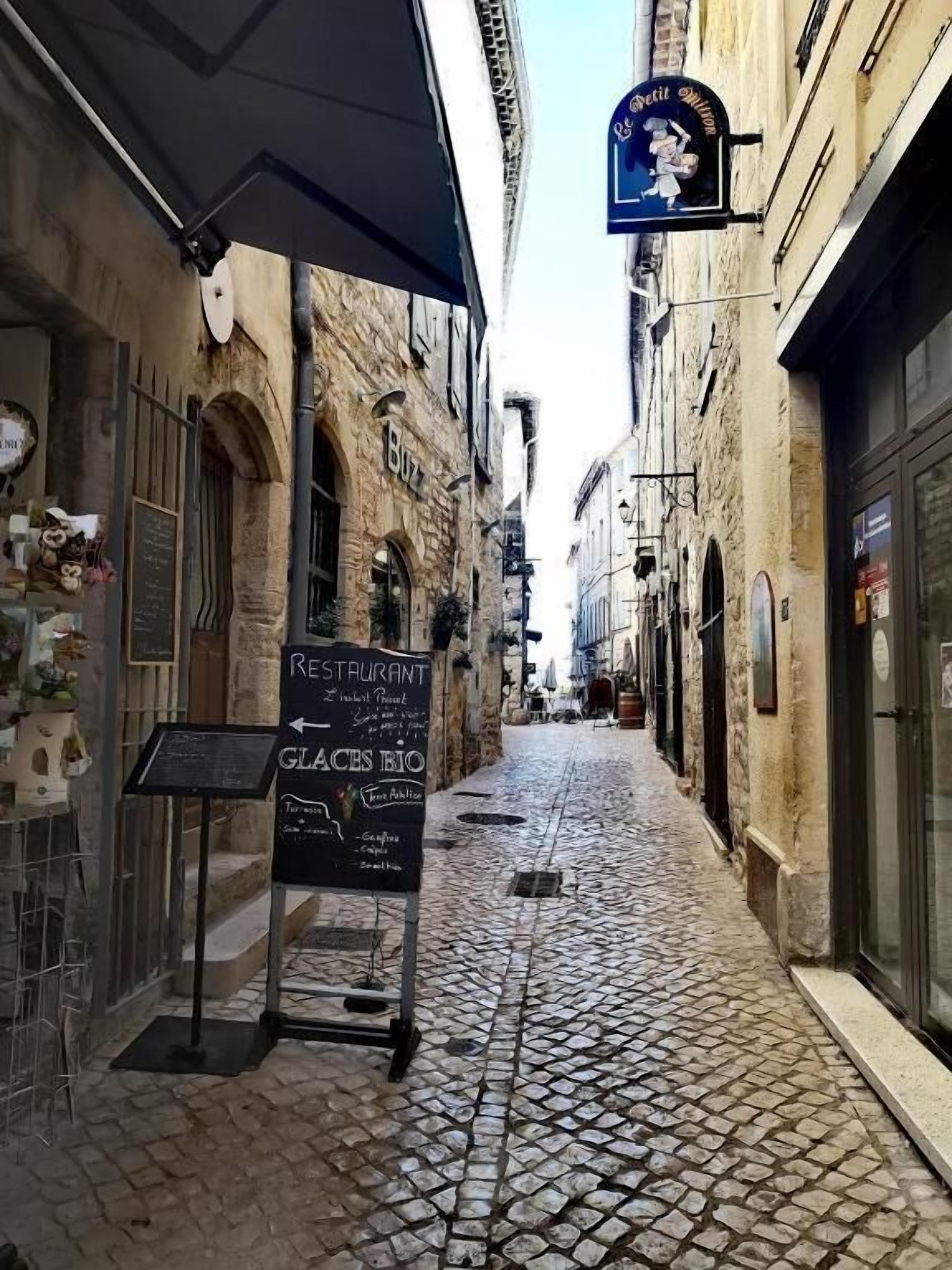 Uma rua de paralelepípedos estreita com o letreiro de um restaurante e uma placa indicando 'GLACES BIO'.