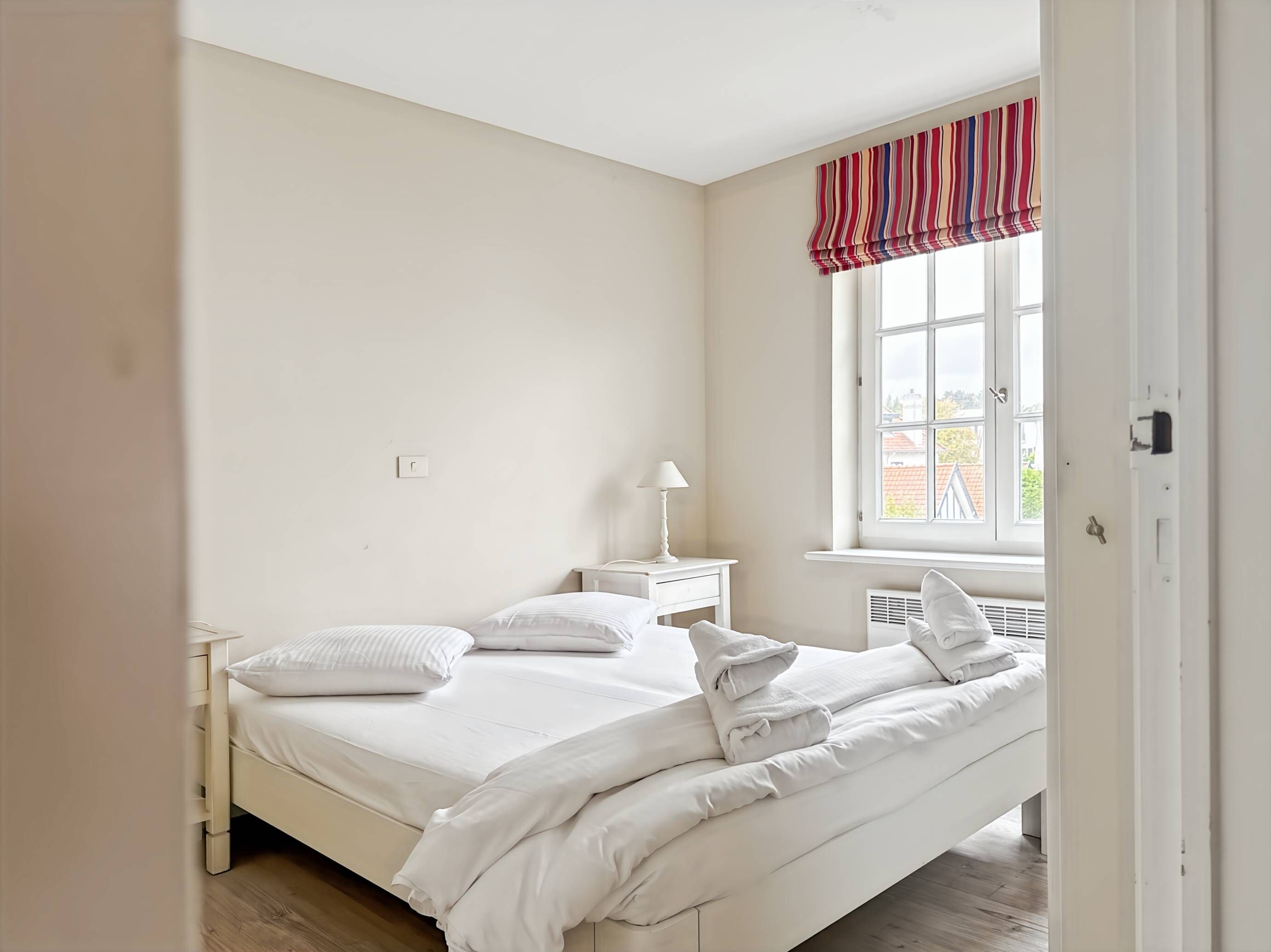 Um quarto com cama, mesas de cabeceira, abajur e janela com cortinas.