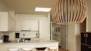 Uma cozinha moderna com armários brancos, uma ilha central com banquetas, uma máquina de café e uma luminária pendente.