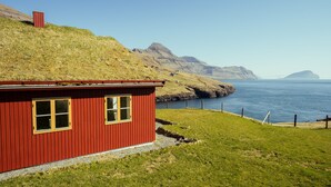 Um edifício vermelho com duas janelas, situado numa colina gramada com vista para um corpo de água e montanhas ao longe.
