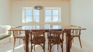 Uma área de refeições com uma mesa e cadeiras de madeira, uma poltrona branca e fofa, e uma janela com vista para o céu.