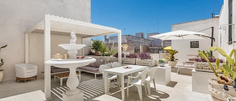 Um terraço no telhado com móveis brancos, uma pérgula branca e plantas em vasos.
