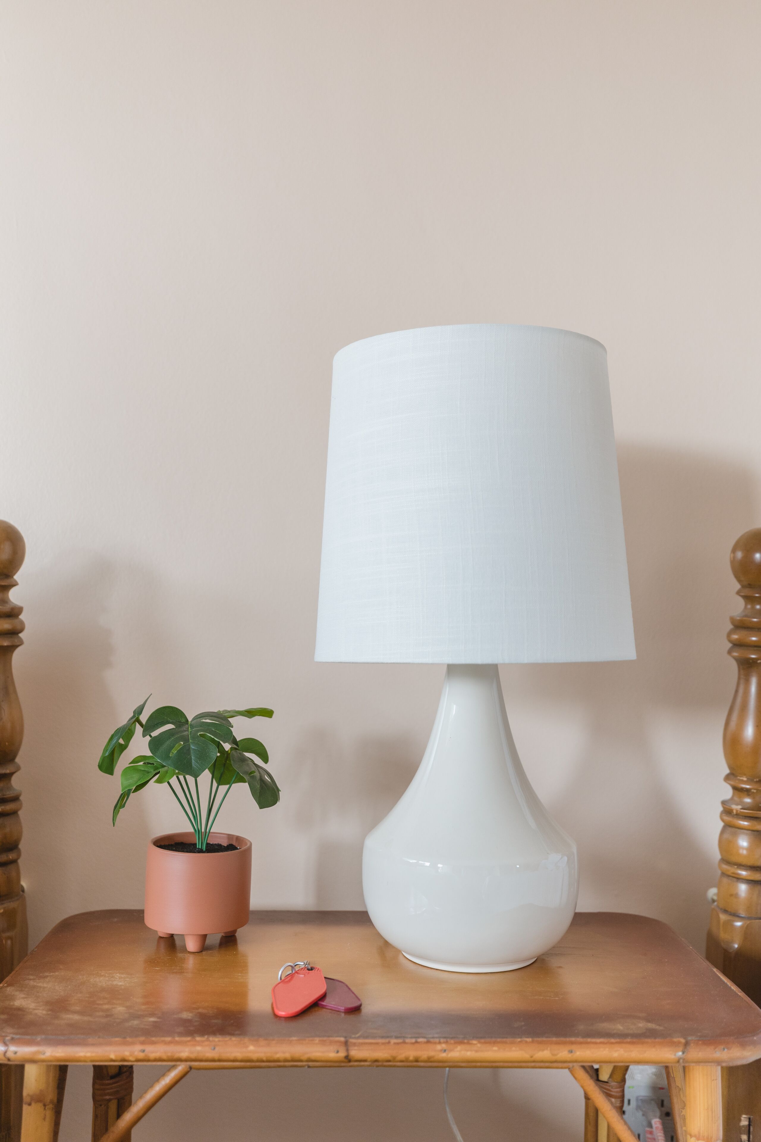 Uma mesa com uma luminária branca, um vaso rosa com uma planta e um chaveiro rosa.