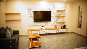 Uma sala de estar moderna com uma TV de tela plana fixada em uma parede de mármore, prateleiras de madeira, uma cafeteira e uma pequena mesa de madeira.