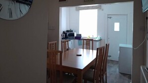 Uma sala de jantar com uma mesa e cadeiras de madeira, uma cozinha ao fundo com eletrodomésticos, e um relógio na parede.
