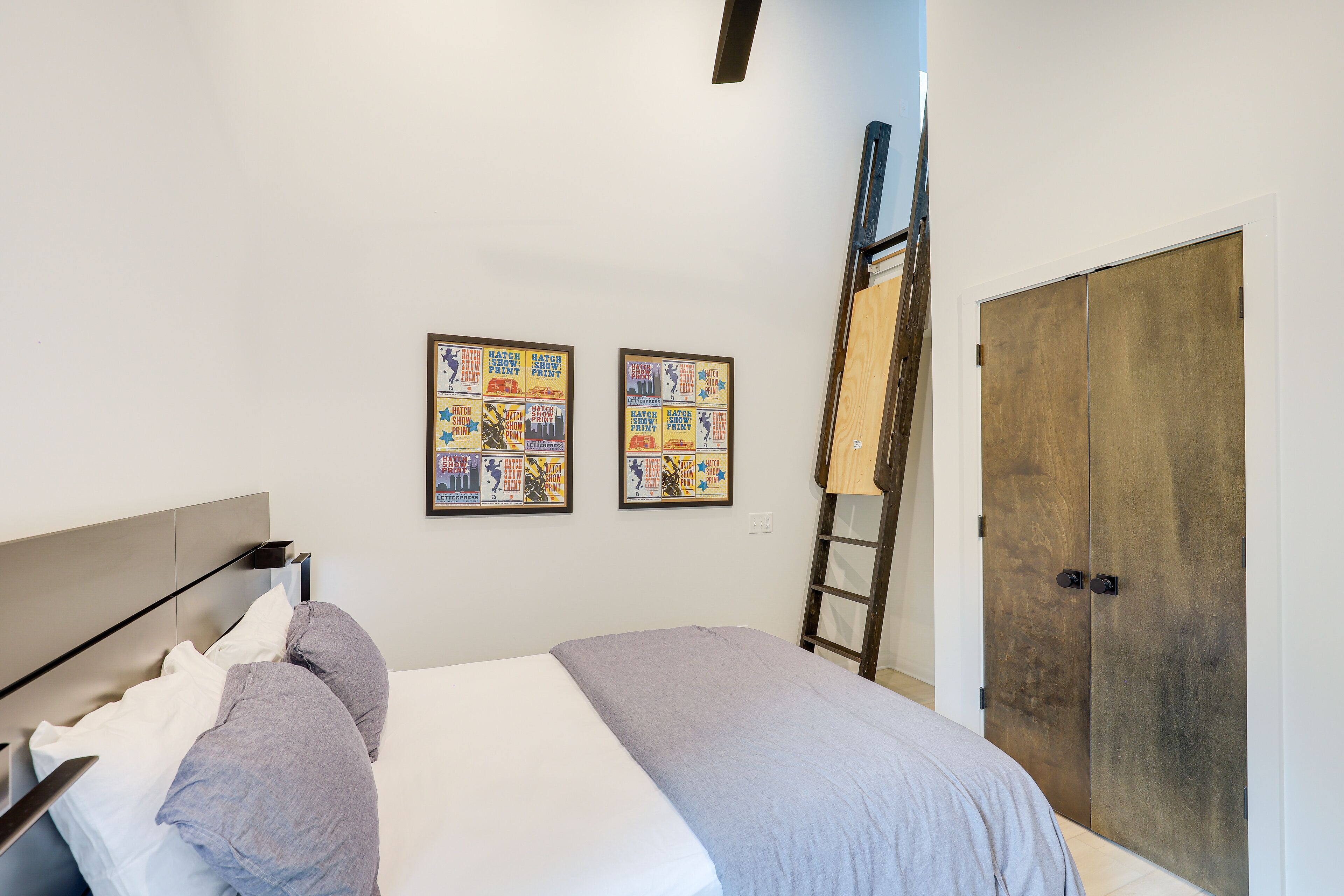 Quarto com cama, almofadas, escada e dois cartazes emoldurados na parede.