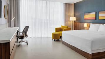 Quarto de hotel com cama, escrivaninha, cadeira, um pequeno banquinho e uma janela grande com cortinas.