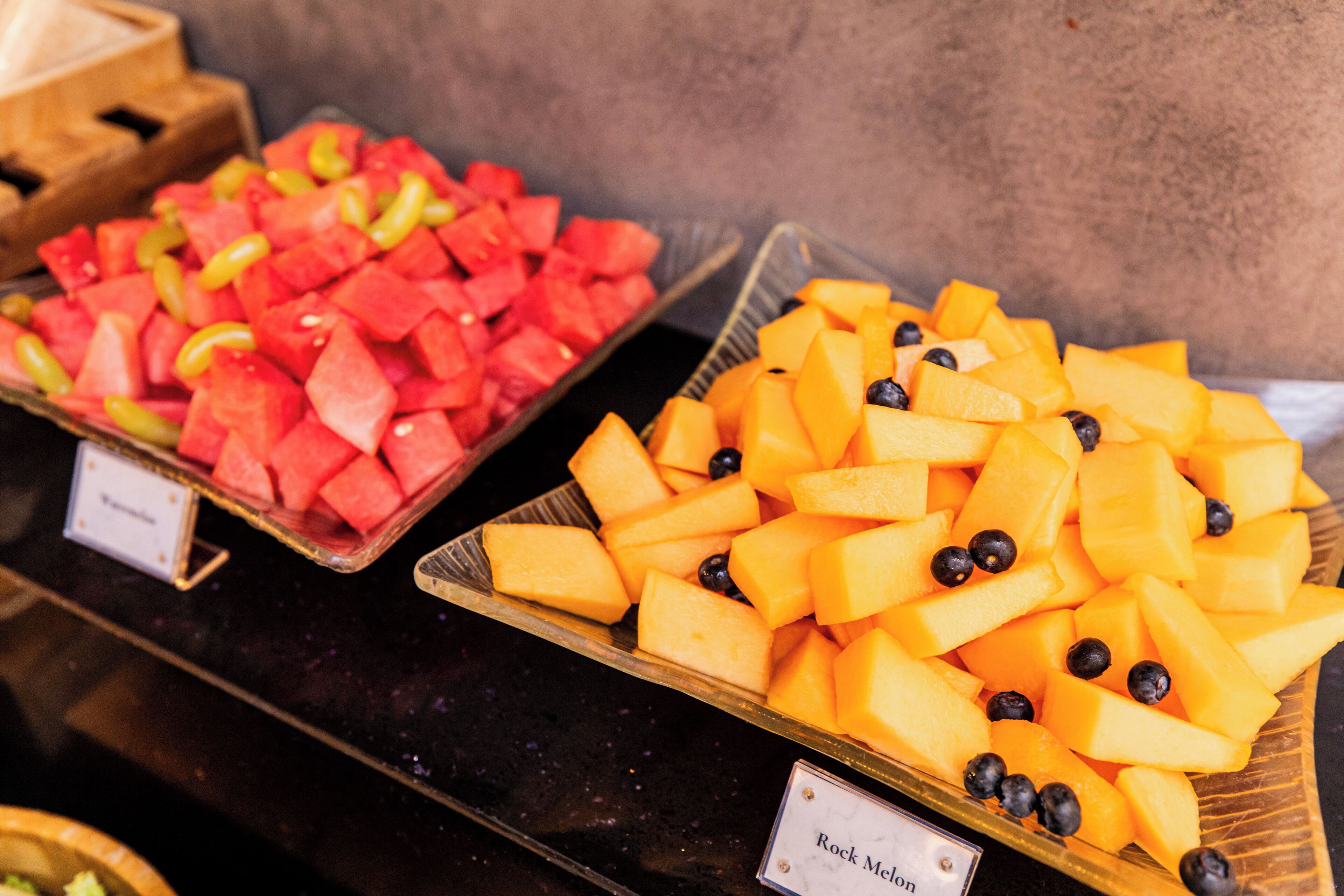 Duas travessas de frutas com os dizeres 'Melancia' e 'Melão', sobre um balcão de mármore.