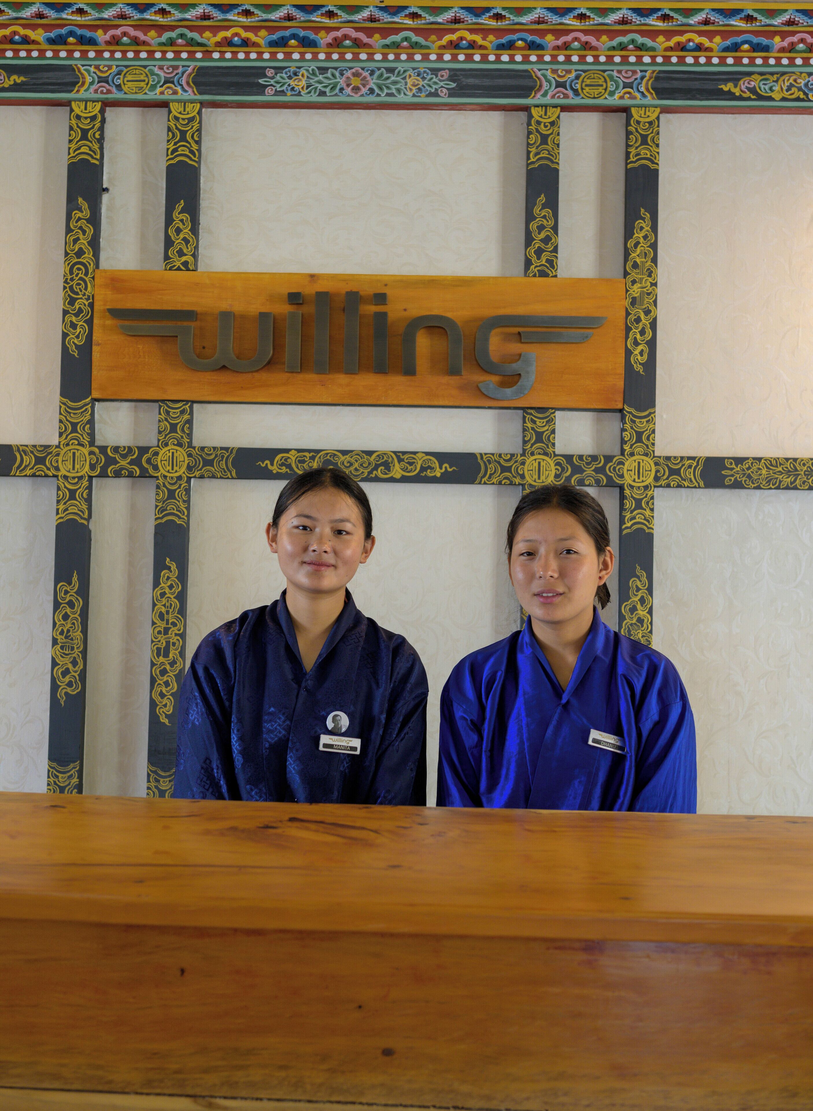 Duas pessoas atrás de um balcão de recepção com uma placa que diz 'willing'.