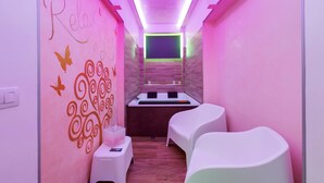 Sala de spa com mesa de massagem, duas cadeiras brancas, parede com mural 'Relax in Life' e pequena mesa com telefone e um recipiente.