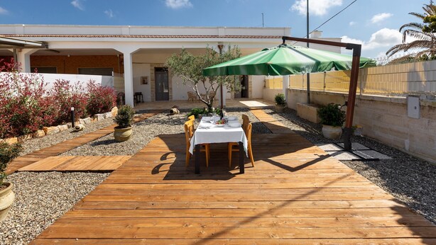 Um deck de madeira com mesa e cadeiras, um guarda-sol verde e um edifício ao fundo.
