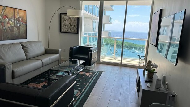 Uma sala de estar moderna com um sofá cinza, uma mesa de centro preta e vista para o oceano através de amplas portas de correr.