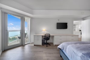 Quarto moderno com uma cama grande, uma escrivaninha com cadeira, TV fixada na parede e porta de vidro deslizante que dá para uma varanda com vista.