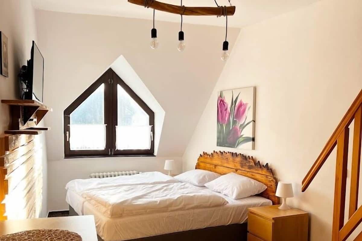 Quarto com teto inclinado, cama com cabeceira de madeira, mesa de cabeceira, luminária, quadro, janela e escada.