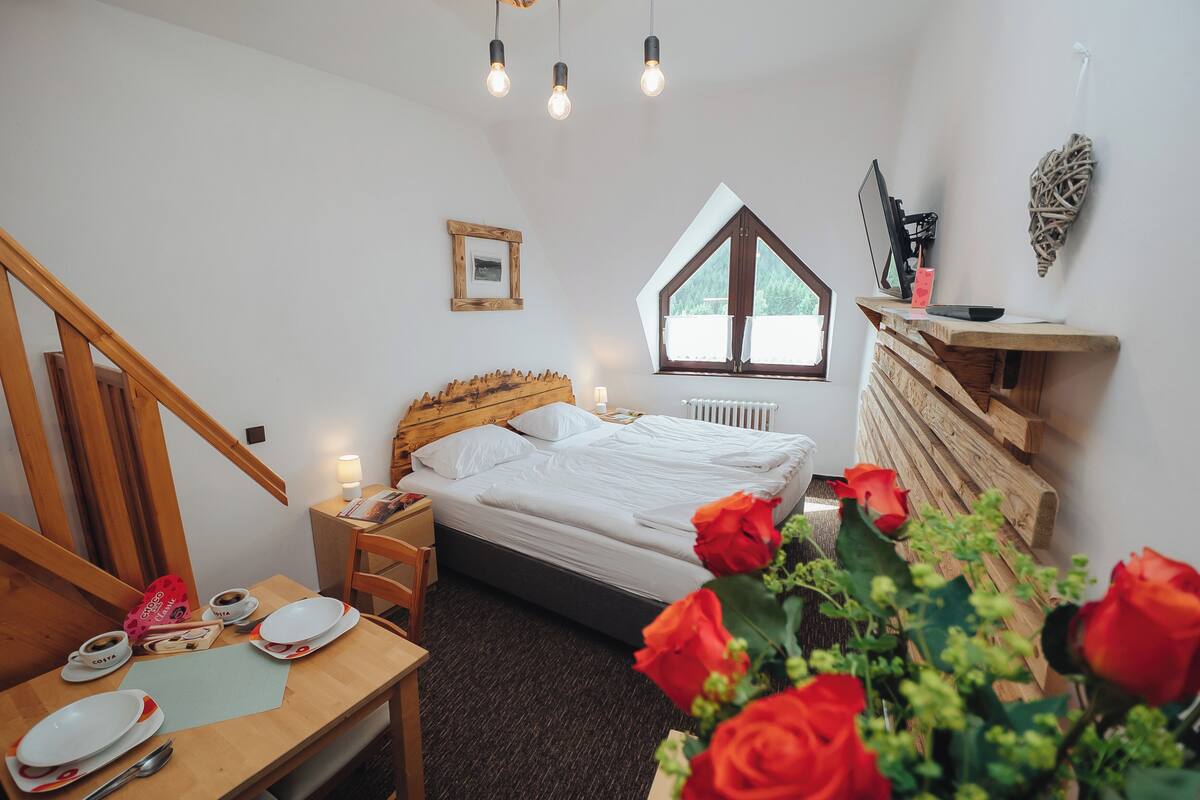 Quarto com cama, cabeceira de madeira, mesa de cabeceira com abajur, janela e pequena estante com objetos decorativos.