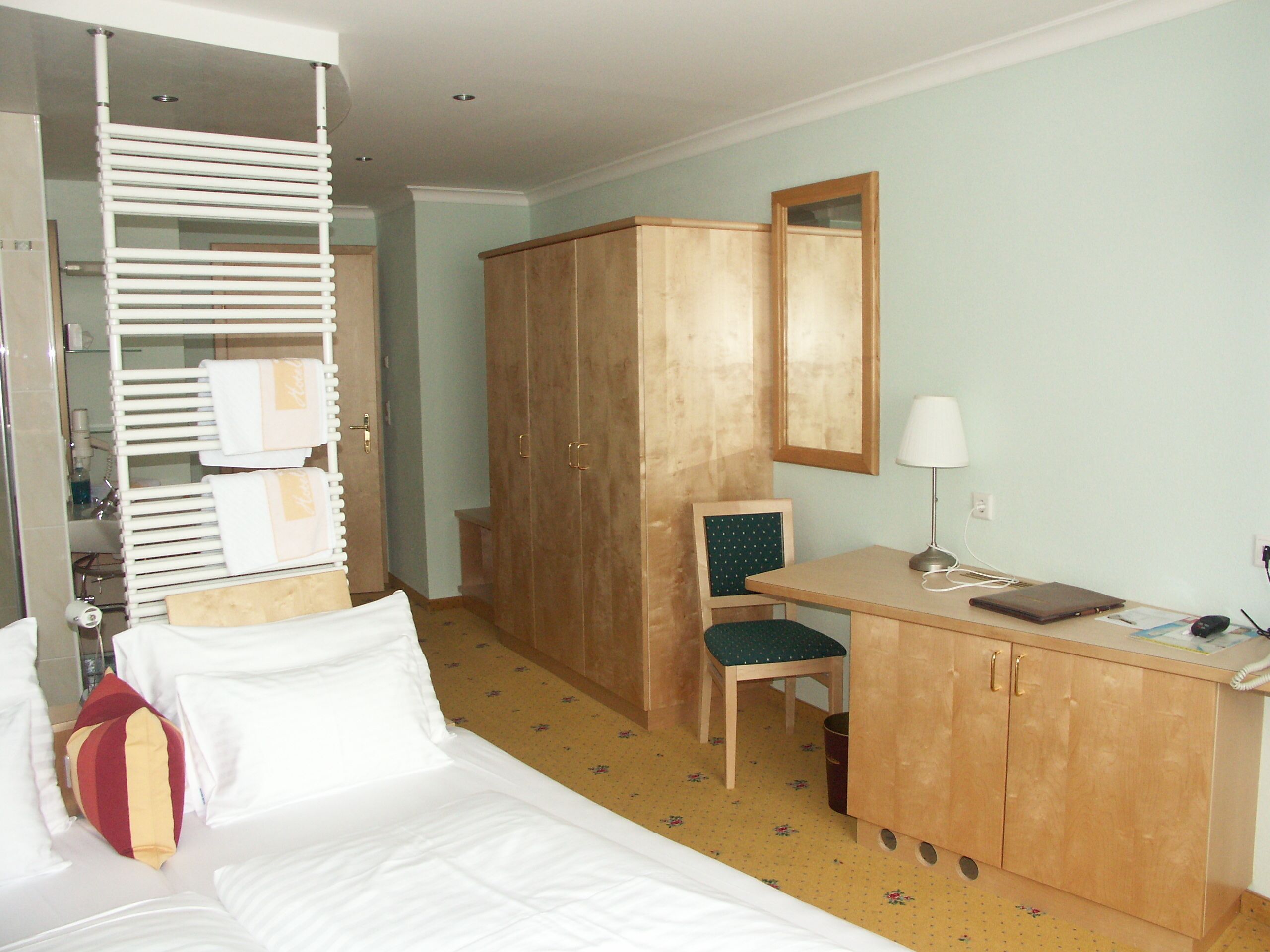 Quarto de hotel com cama, escrivaninha, cadeira, guarda-roupa e espelho.