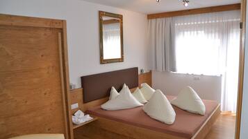 Quarto com cabeceira de madeira, cama com almofadas brancas, mesa de cabeceira de madeira, espelho e janela com cortinas.
