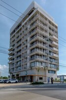 Edifício de vários andares com varandas, entrada no nível da rua e movimentação de carros e pedestres visíveis.
