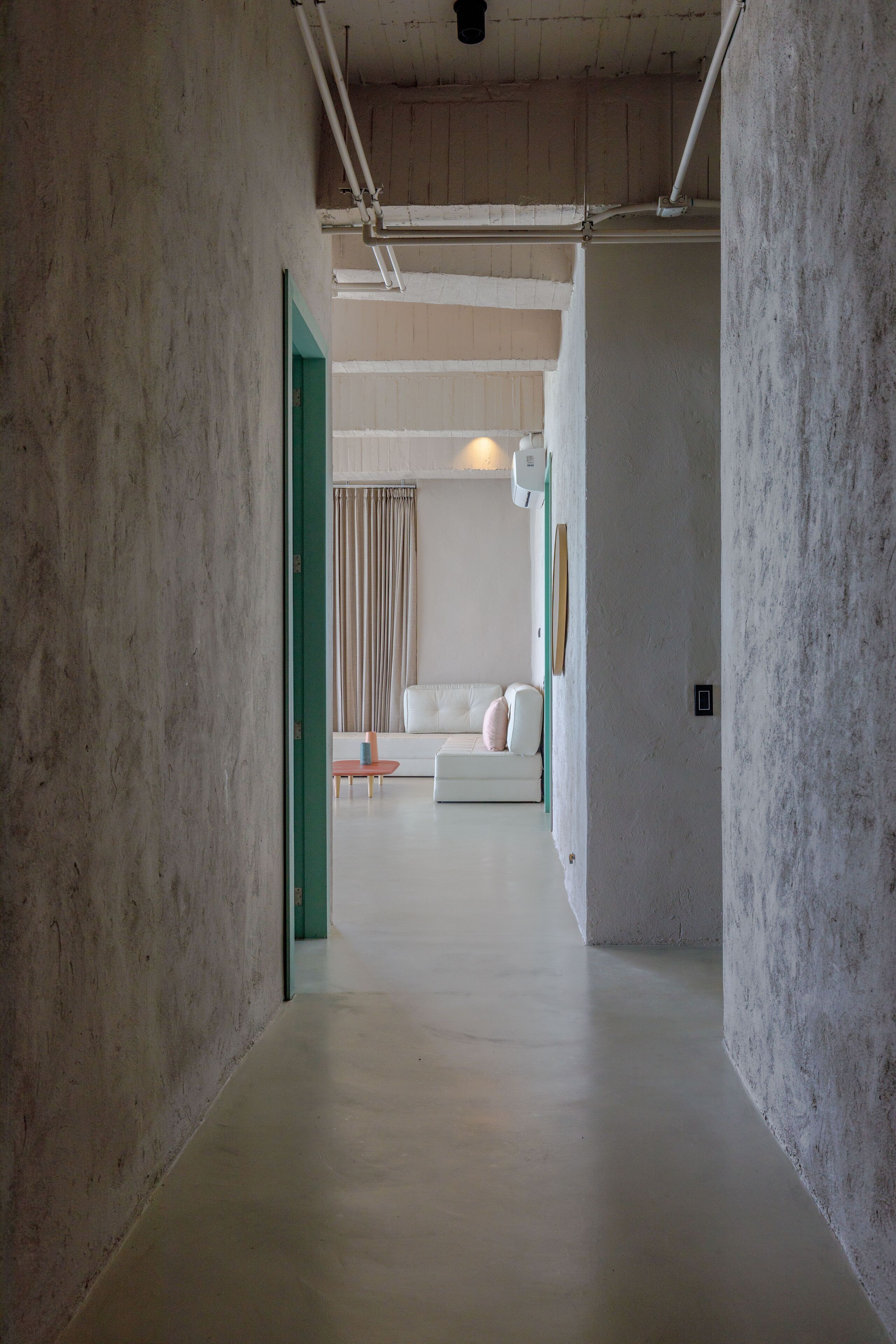 Corredor que leva a uma sala de estar com sofá branco e uma mesinha, paredes de concreto e teto com tubulações aparentes.
