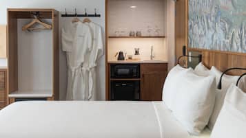 Quarto de hotel com cama, guarda-roupa de madeira, suporte para roupões, micro-ondas, máquina de café e pia.
