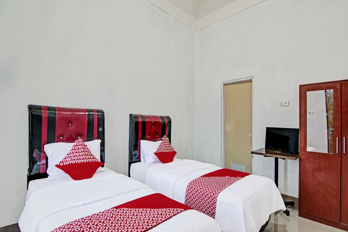 Quarto de hotel com duas camas, cada uma com roupa de cama vermelha e branca, uma televisão sobre uma mesinha e um guarda-roupa de madeira.