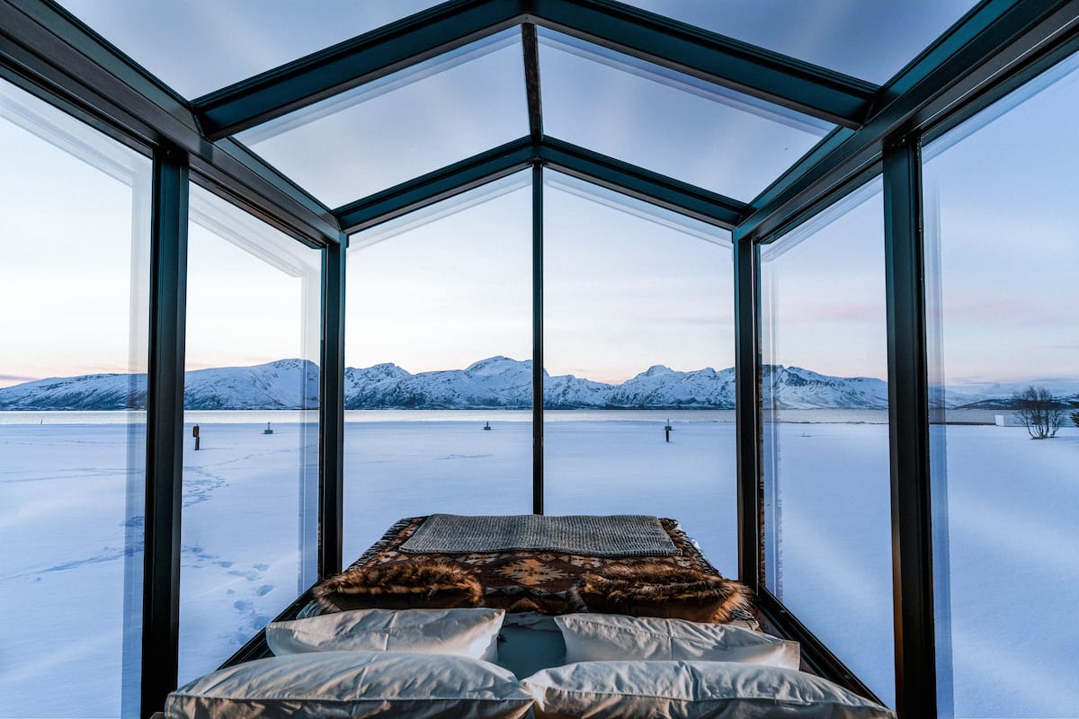 Quarto com teto de vidro, cama com roupa de cama branca e marrom, e vista para uma paisagem nevada.