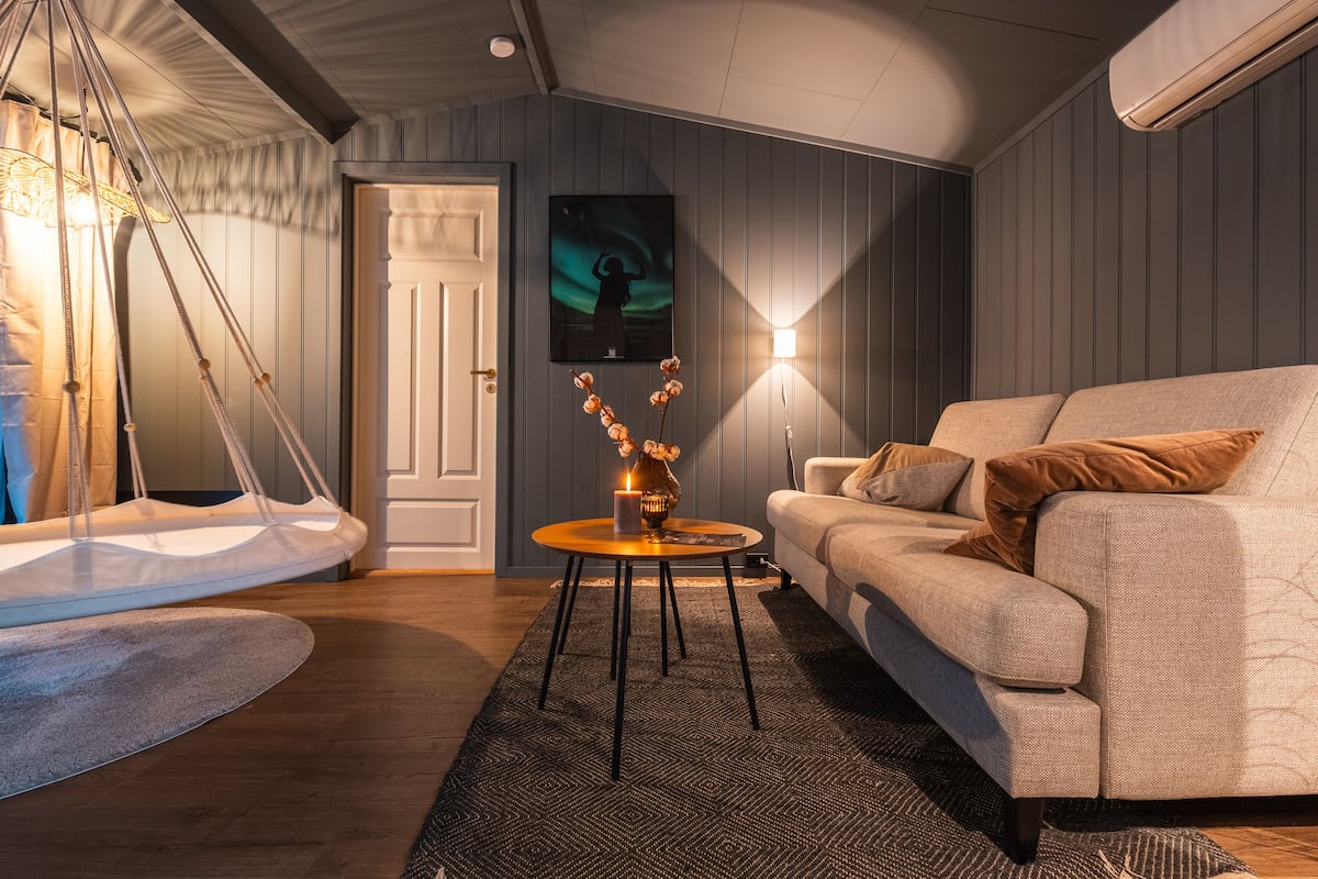 Uma sala de estar acolhedora com uma cadeira suspensa, um sofá, uma mesinha com uma vela e uma TV fixada na parede.
