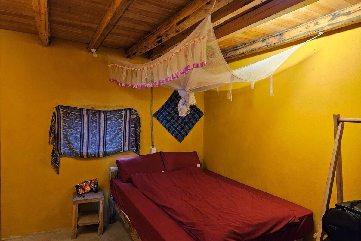 Quarto com cama, uma mesinha, um banco, mosquiteiro e parede com tecido estampado.