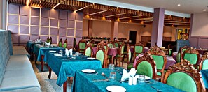 Uma área de refeições com várias mesas preparadas para hóspedes, com cadeiras estofadas em verde e toalhas de mesa azuis.