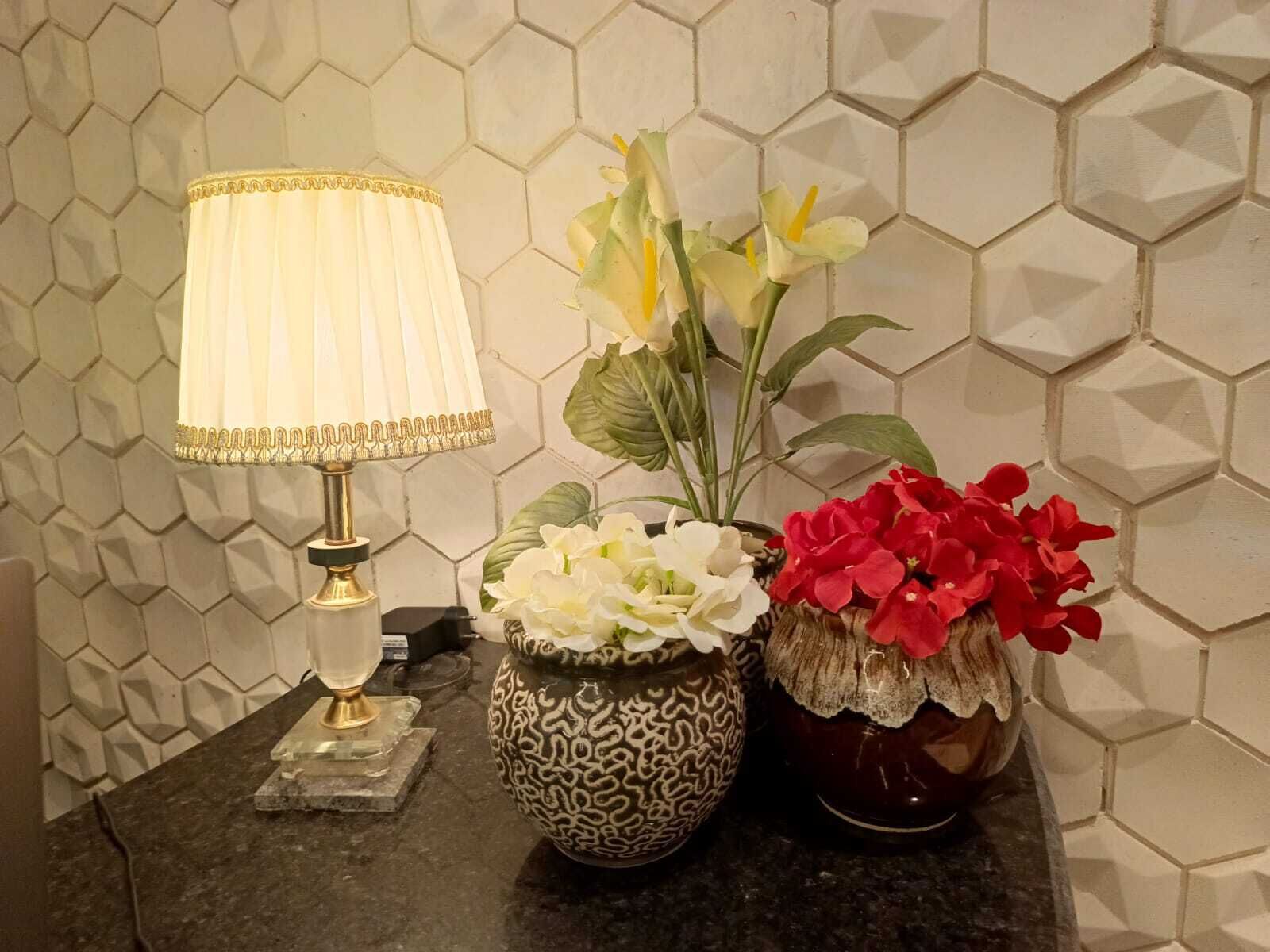 Uma mesa com uma lâmpada, dois vasos com flores e um painel de azulejos hexagonais.